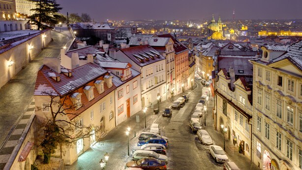 Uma rua de paralelepípedos coberta de neve, com edifícios históricos, carros estacionados e uma imponente torre de igreja ao fundo.