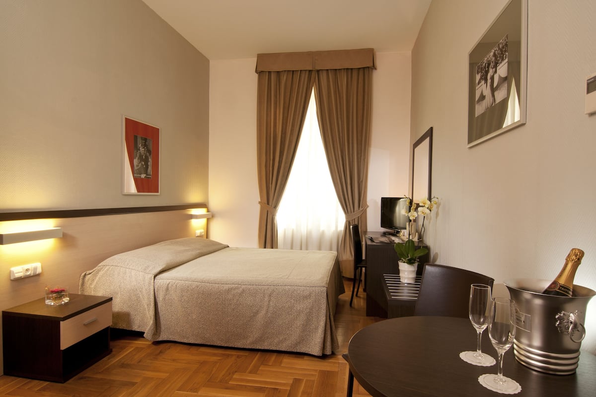 Quarto de hotel com cama, mesa de cabeceira, televisão, escrivaninha com uma planta e mesa de jantar com dois copos e uma garrafa de champanhe.