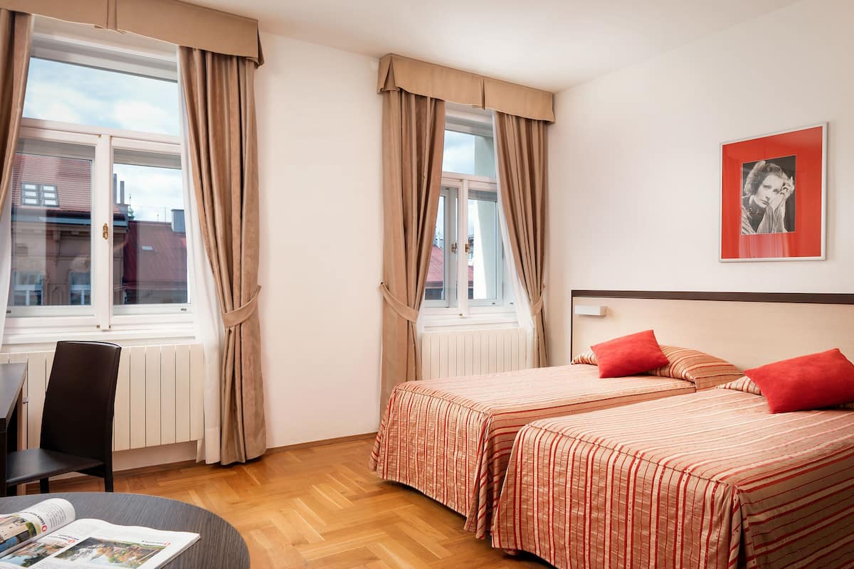 Quarto de hotel com duas camas, uma cadeira, uma mesa e uma janela com cortinas.