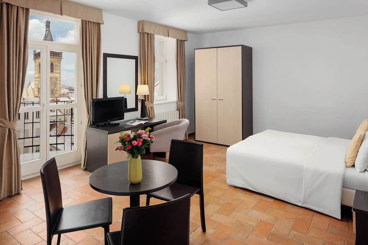 Quarto de hotel com cama, escrivaninha com televisão, cadeira, mesa com um vaso de flores e vista para a cidade através de amplas janelas.