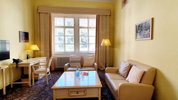 Quarto de hotel com sofá, mesa de centro, televisão, escrivaninha com abajur e janela com cortinas.