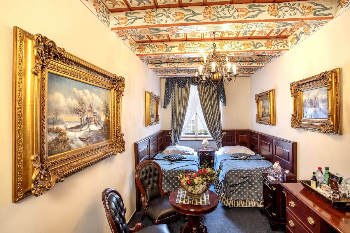 Quarto de hotel com duas camas, um candelabro, teto ricamente decorado e obras de arte emolduradas.