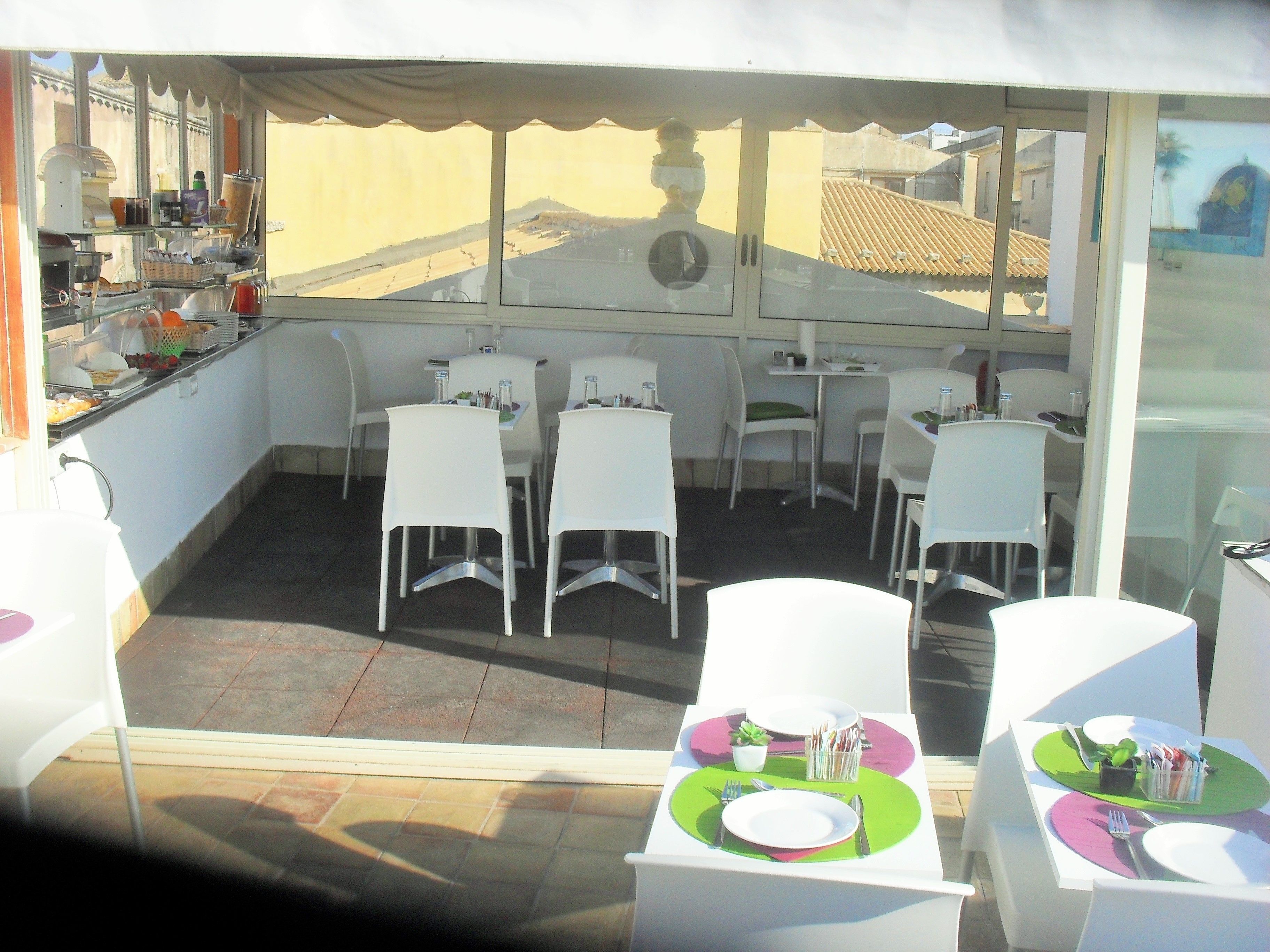 Uma área de refeições ao ar livre coberta, com cadeiras e mesas brancas, um balcão de buffet e vista para edifícios ao fundo.