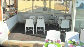Uma área de refeições ao ar livre coberta, com cadeiras e mesas brancas, um balcão de buffet e vista para edifícios ao fundo.