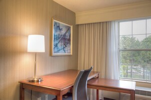 Quarto de hotel com secretária de madeira, uma cadeira, abajur, quadro na parede e janela com cortinas.