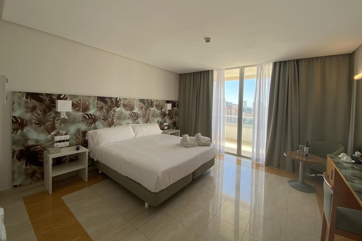 Quarto de hotel moderno com uma cama grande, uma mesinha de cabeceira, uma escrivaninha com cadeira, e vista para a cidade pela janela.