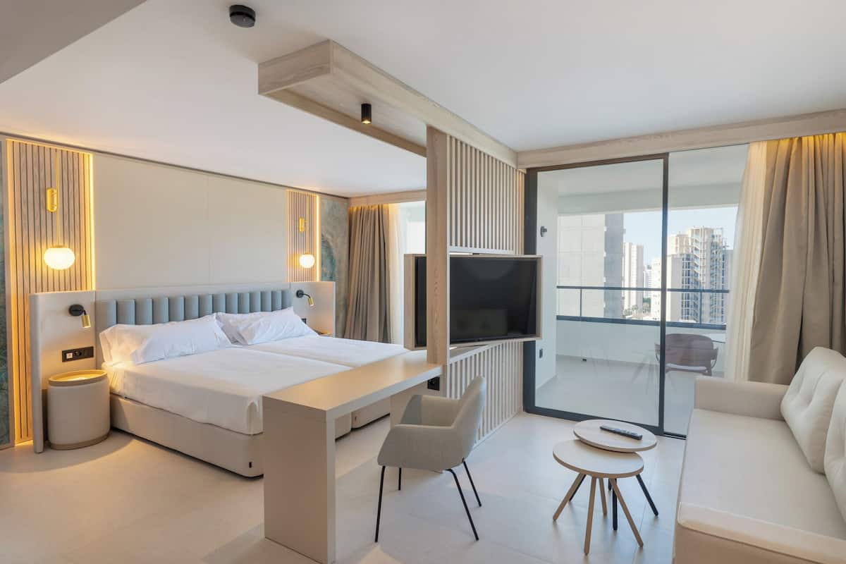 Quarto de hotel moderno com uma cama grande, televisão, sofá e varanda com vista para a cidade.