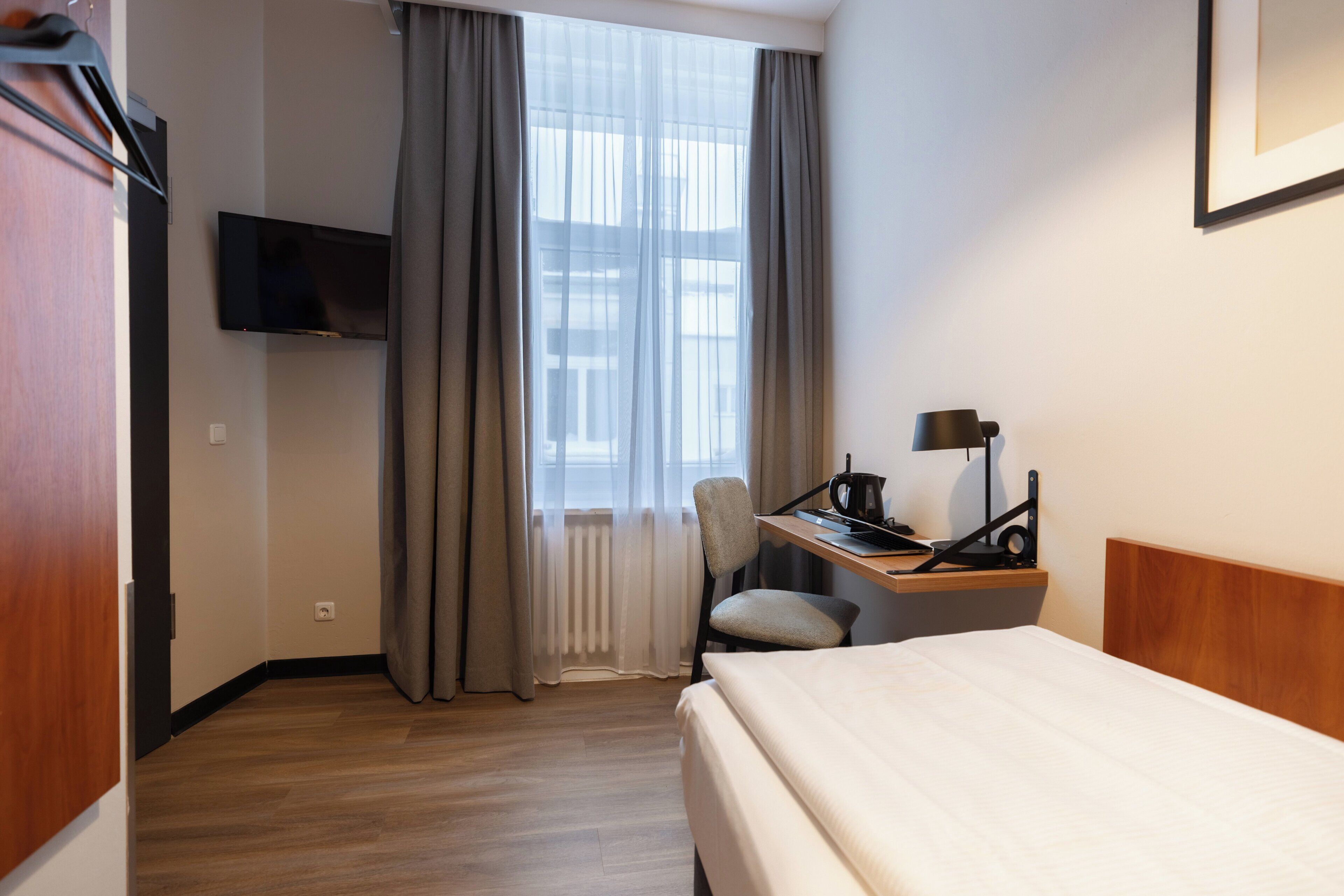 Quarto de hotel com cama, escrivaninha com cadeira, televisão na parede e janela com cortinas.