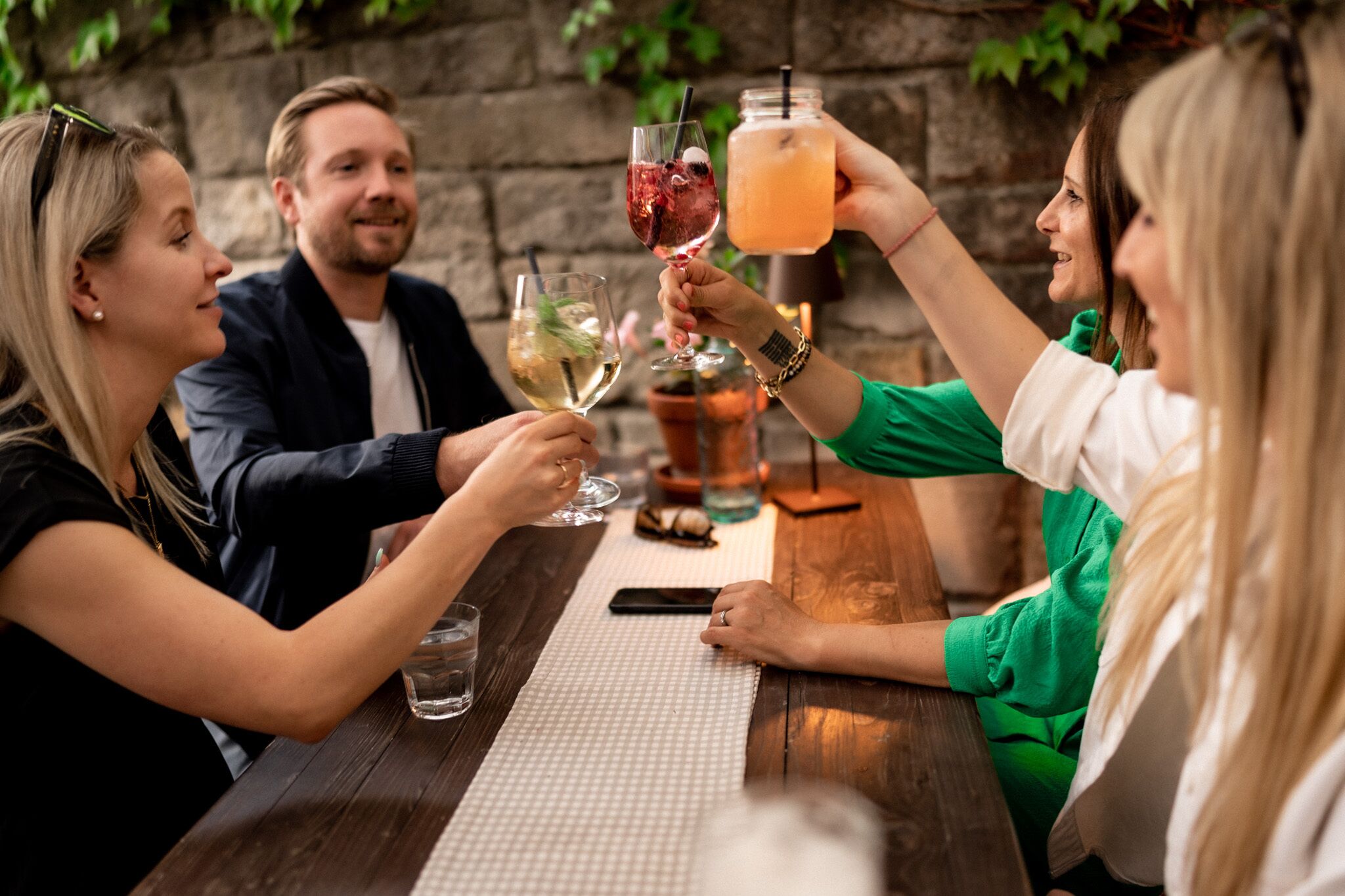 Um grupo de pessoas brindando com bebidas em uma mesa de madeira, com um muro de pedra ao fundo.