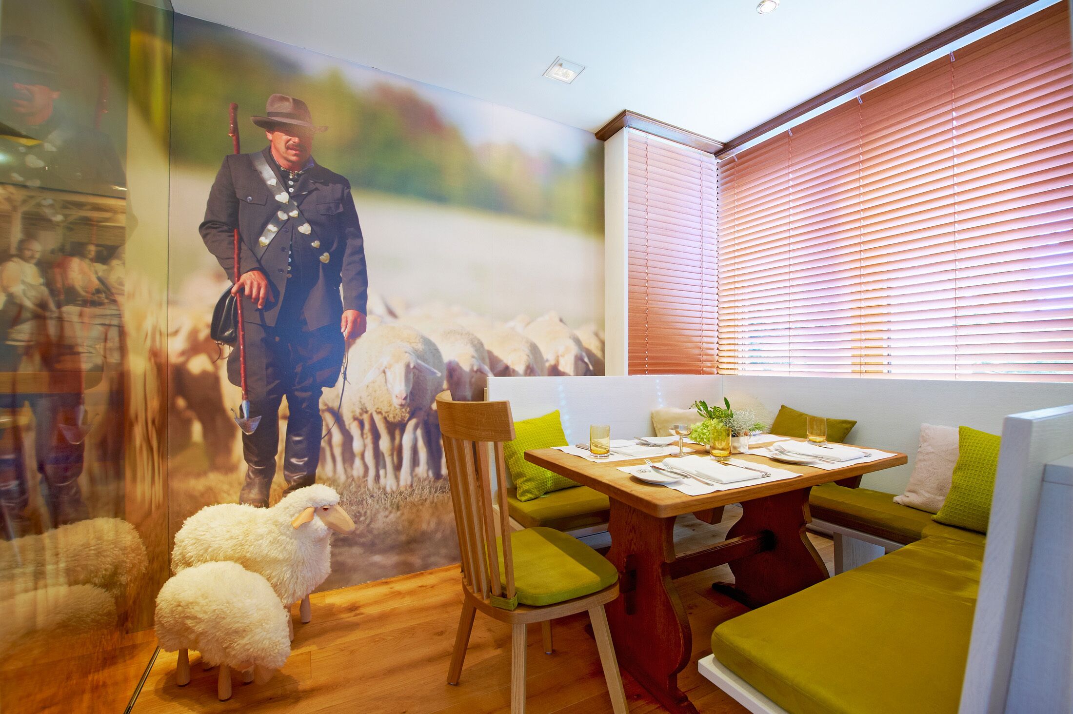 Uma área de jantar com mesa de madeira, assentos acolchoados verdes e um mural de um homem com ovelhas.