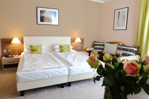Quarto de hotel com cama de casal, duas mesas de cabeceira, duas luminárias, um criado-mudo, uma cadeira e um vaso com flores.
