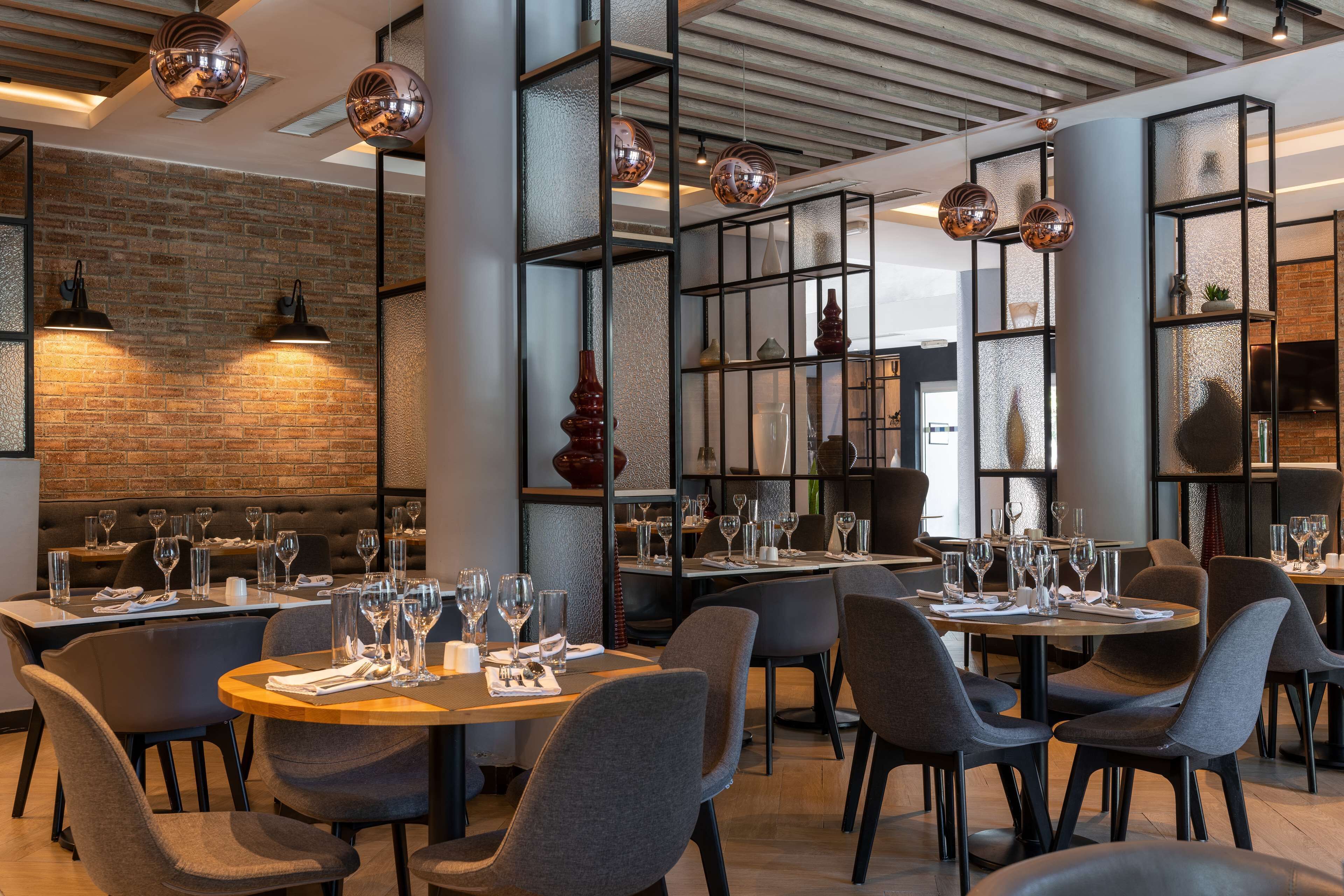 Restaurante com mesas redondas, cadeiras cinzentas e parede de tijolos.