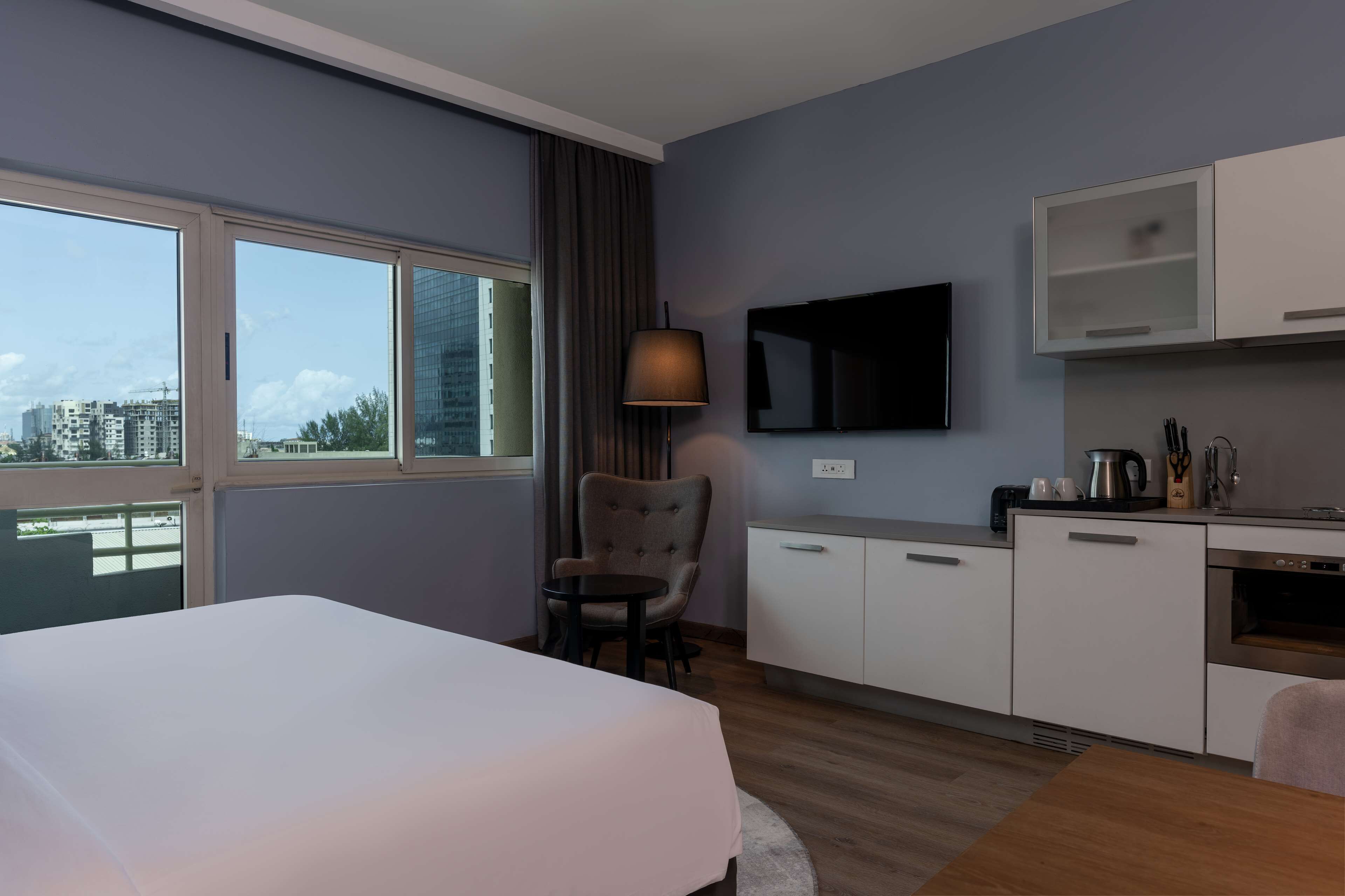 Quarto de hotel com cama, escrivaninha, cadeira, televisão e cozinha compacta.
