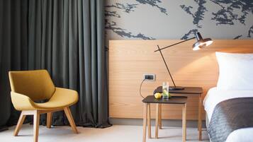 Quarto de hotel com cabeceira de madeira, cama, mesa de cabeceira com abajur, pequena mesa com um copo de água e um limão, e uma cadeira cor mostarda.