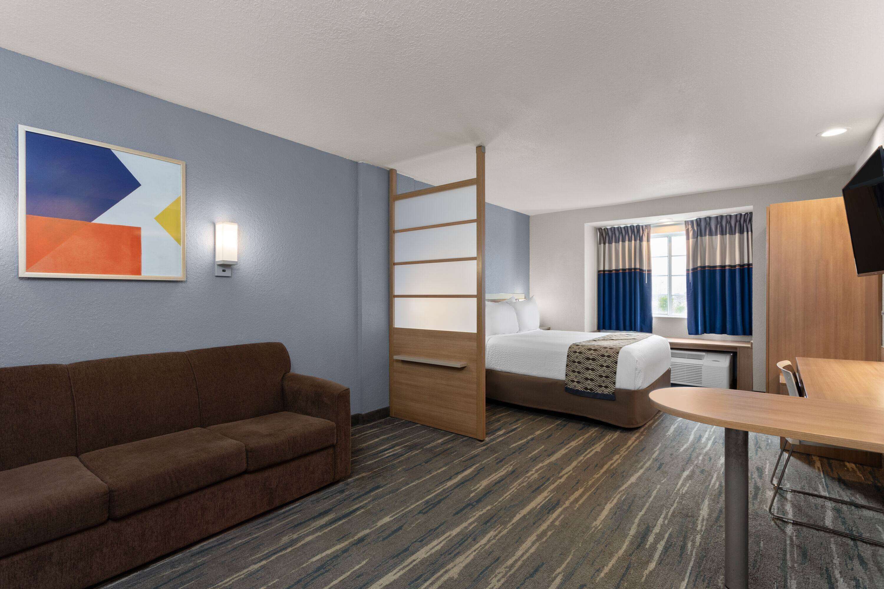Quarto de hotel com sofá marrom, cama com cabeceira branca, escada de madeira, escrivaninha com cadeira e um quadro na parede.