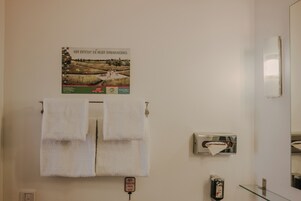 Casa de banho com duas toalhas brancas penduradas num suporte, um dispensador de papel e um aviso a indicar proibição de estacionamento.