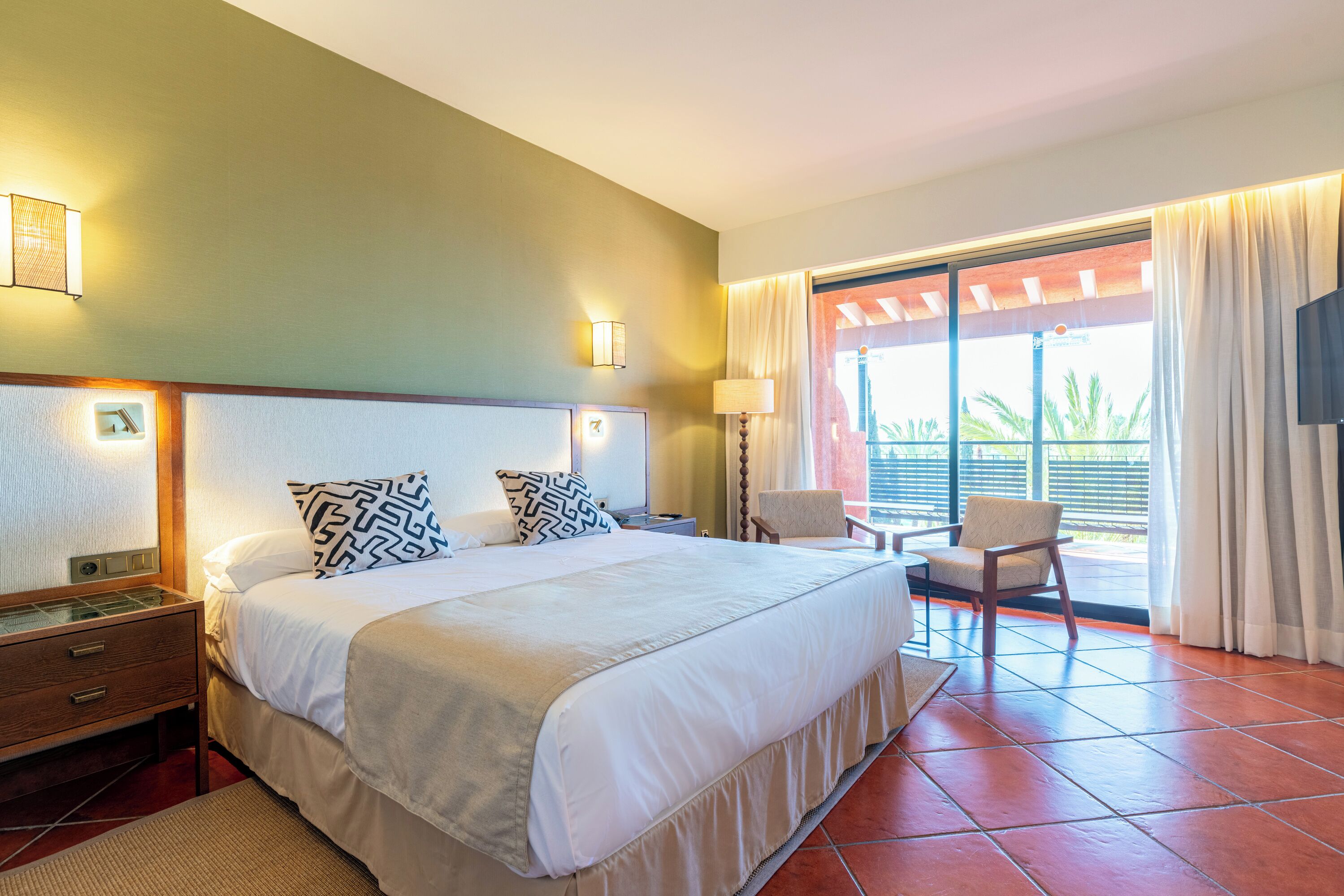 Quarto de hotel com uma cama grande, uma mesinha de cabeceira, uma cadeira e vista para o exterior.