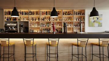 Uma área de bar com balcão de madeira, luminárias pendentes pretas, banquetas de vime e prateleiras com diversas garrafas de bebidas.