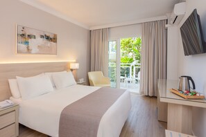 Quarto de hotel com cama, escrivaninha com chaleira, cadeira, televisão e varanda com cortinas.