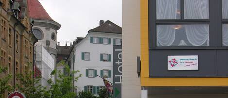 Um hotel com uma placa que diz "Hotel Sonne-Beck" e uma placa com "Top of Thurgau". Há uma estátua de uma pessoa em pernas de pau em frente ao hotel.