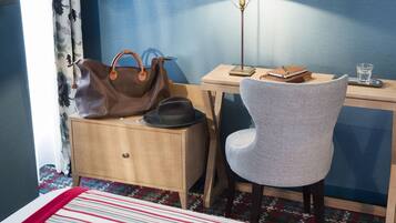 Quarto de hotel bem arrumado, com uma cama, uma escrivaninha com abajur, uma cadeira, uma bolsa, um chapéu, um caderno e um copo de água.