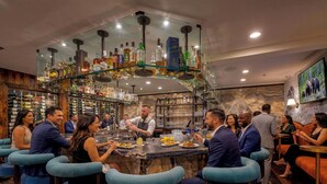 Uma área de bar com mesas redondas, cadeiras estofadas em azul e uma variedade de garrafas de bebidas alcoólicas nas prateleiras.