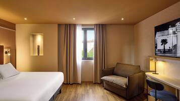 Quarto de hotel com cama, escrivaninha com cadeira, janela com cortinas e um quadro pendurado na parede.