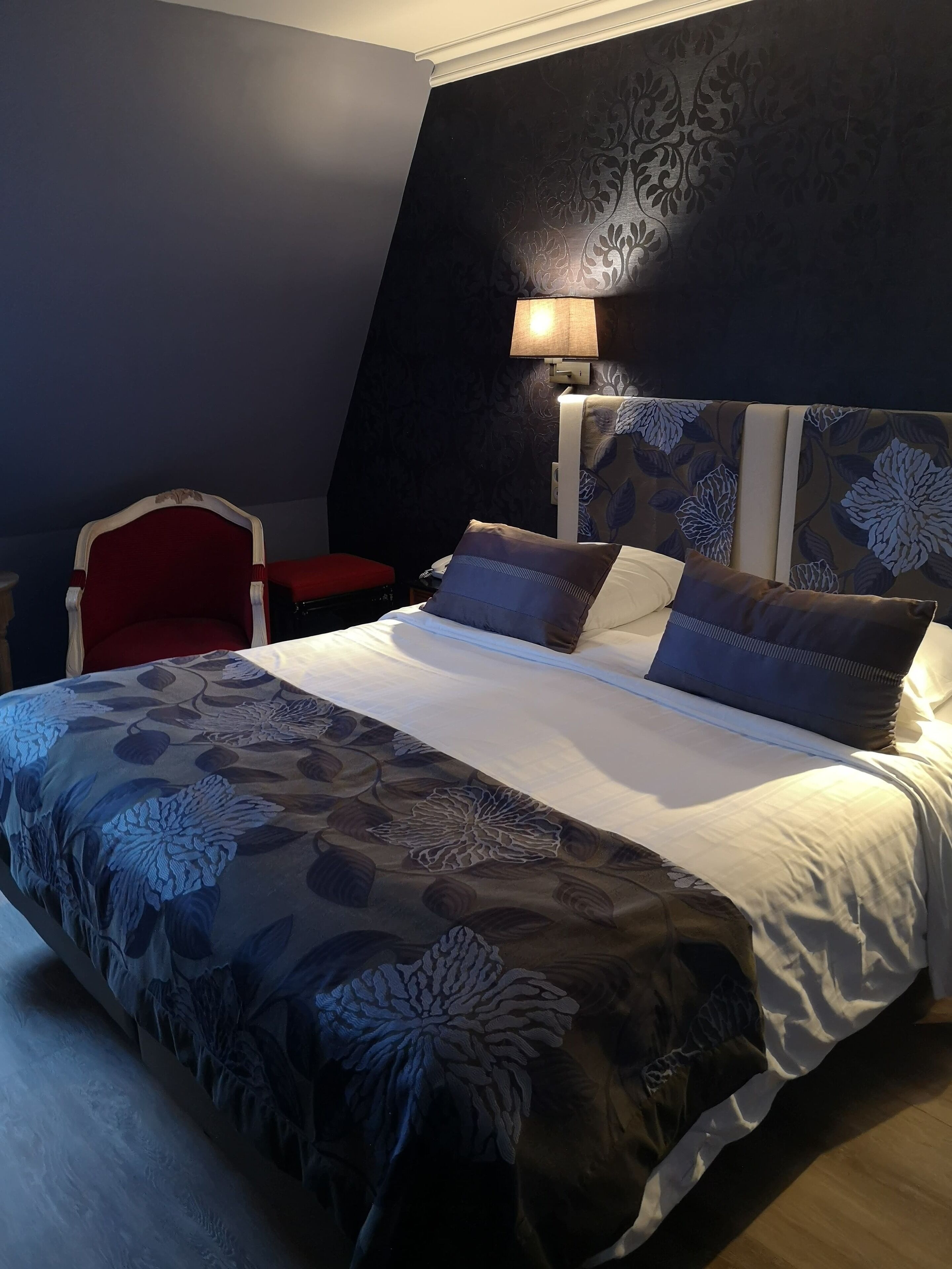 Quarto de hotel com uma cama grande, uma poltrona vermelha, uma mesa de cabeceira e uma parede com padrão floral.