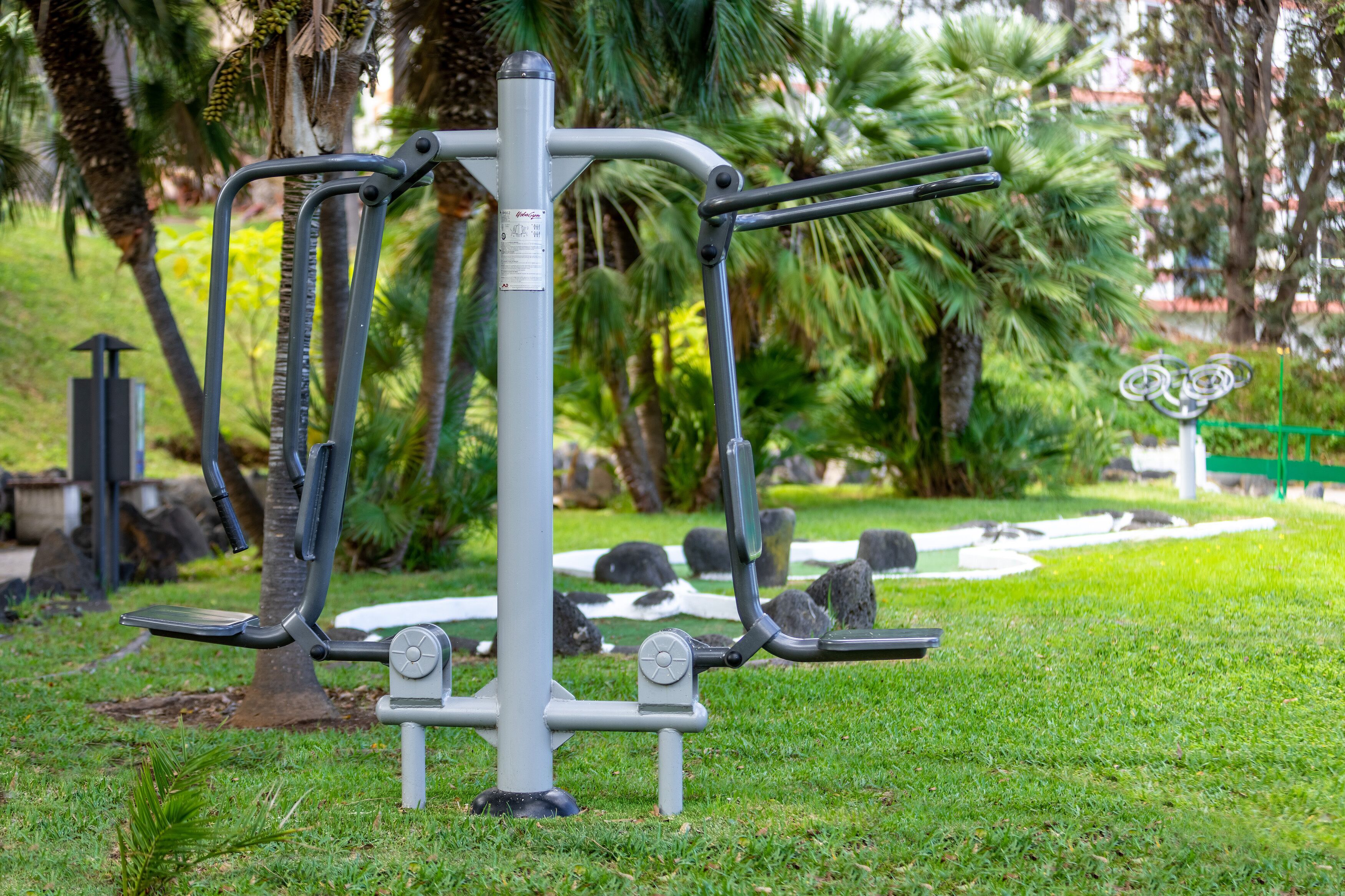 Aparelho de ginástica ao ar livre em um parque com vegetação e um caminho de pedra.