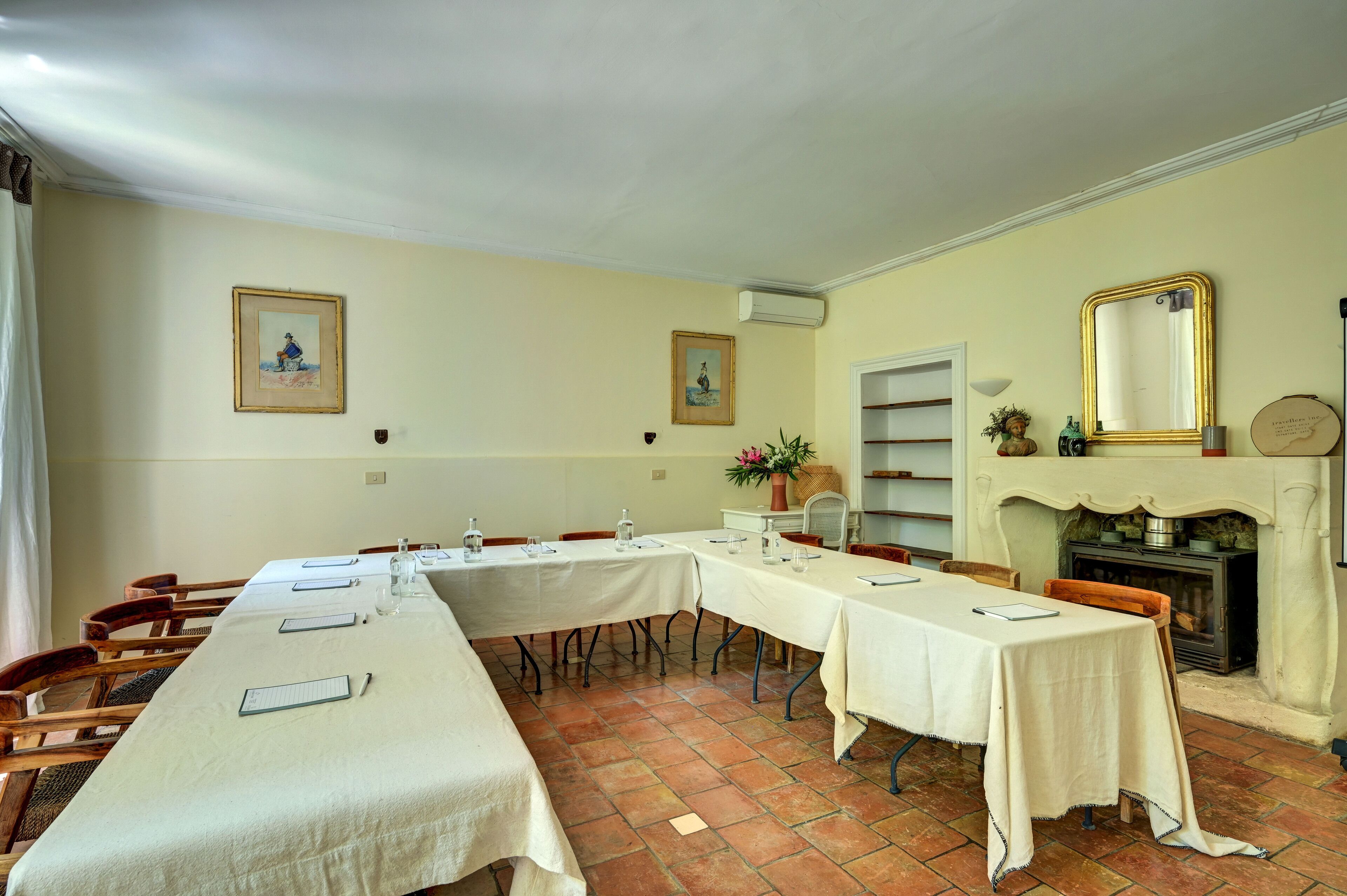 Uma sala de jantar com mesas compridas, cadeiras, lareira e quadros nas paredes.