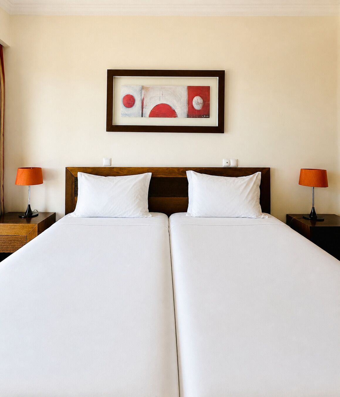 Uma cama de casal com lençóis brancos, duas mesas de cabeceira com luminárias e um quadro abstrato na parede.