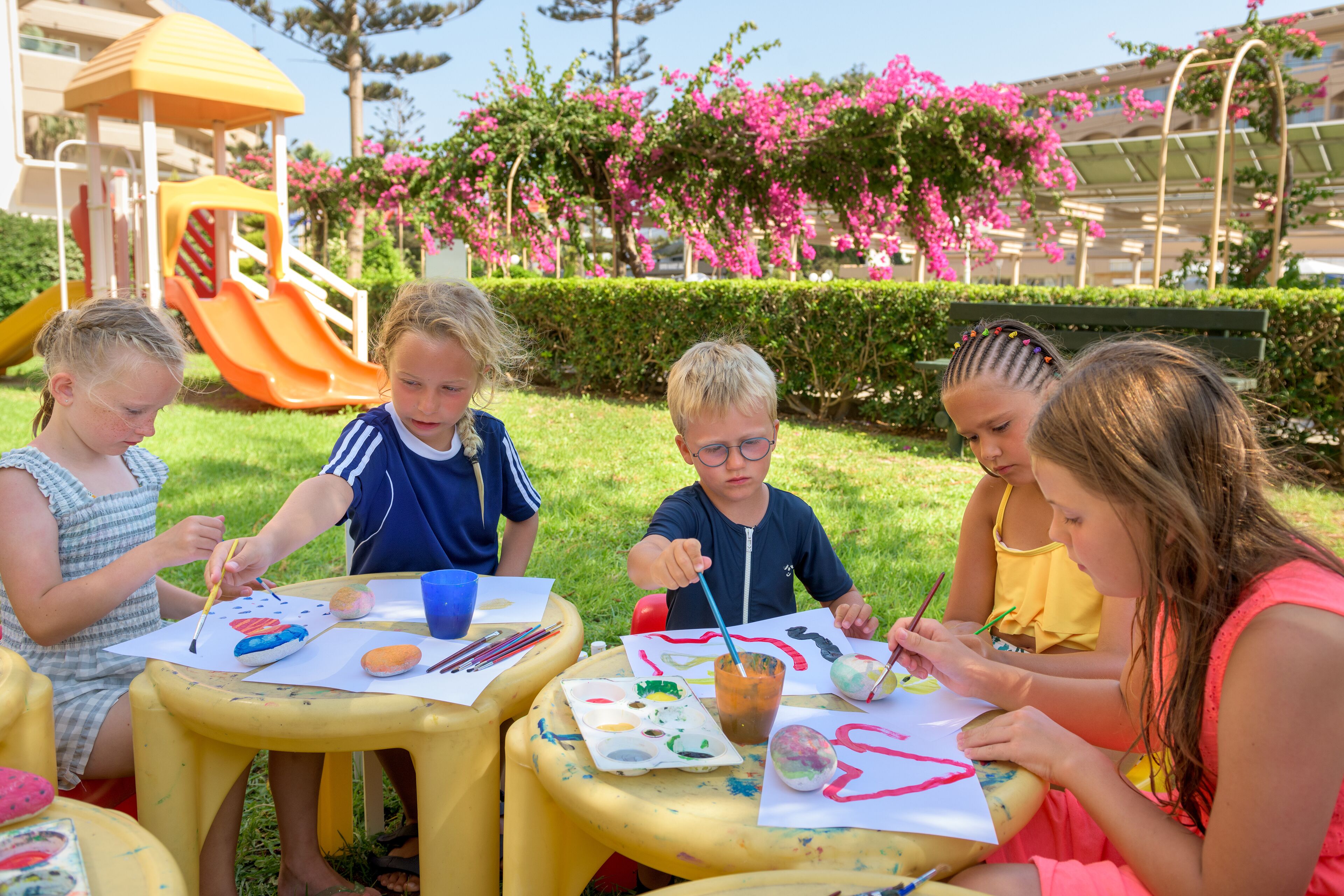 Um grupo de crianças pintando ao ar livre, com um parquinho ao fundo.
