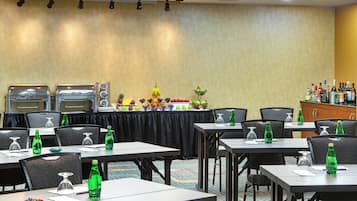 Sala de conferências com mesas, cadeiras e mesa de buffet com frutas e bebidas.