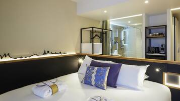 Quarto de hotel com uma cama grande, um espelho, um banheiro visível através de uma porta aberta e uma prateleira com artigos de higiene pessoal.