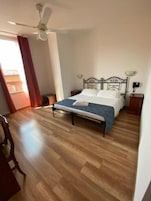 Quarto com cama, ventoinha no teto, piso de madeira, mesa de cabeceira e janela com cortinas.