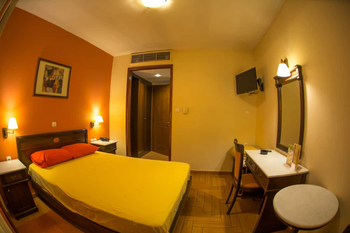 Quarto de hotel com cama, mesa de cabeceira, luminária, cadeira, escrivaninha, espelho e um quadro na parede.