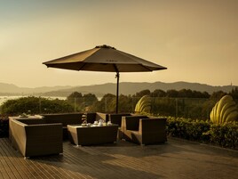 Um terraço com móveis de vime, uma mesa com uma chaleira e um grande guarda-sol.