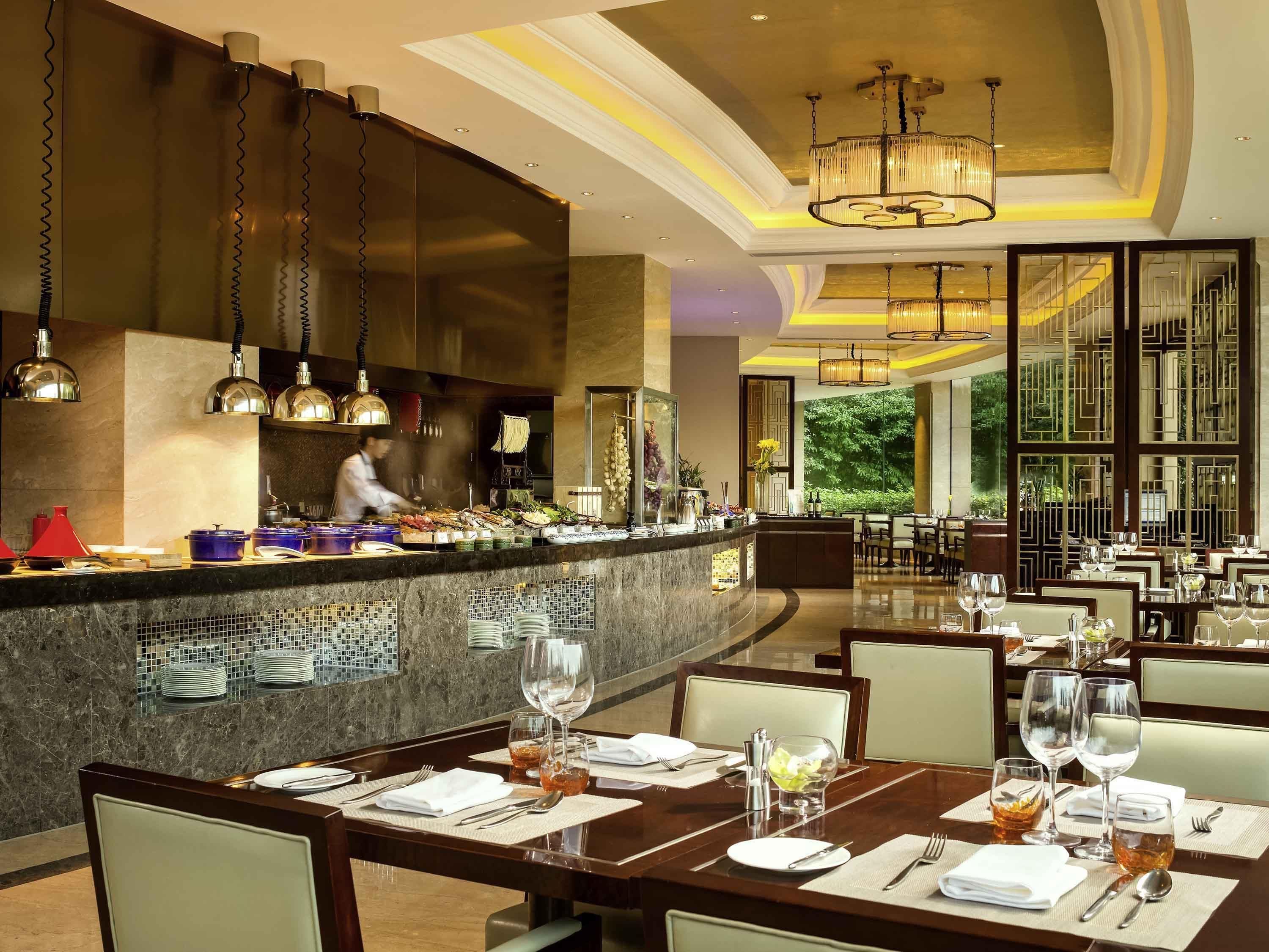 Um restaurante moderno com um bar central, mesas de jantar dispostas com copos de vinho e talheres, e um chef preparando alimentos ao fundo.