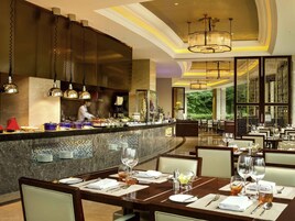Um restaurante moderno com um bar central, mesas de jantar dispostas com copos de vinho e talheres, e um chef preparando alimentos ao fundo.