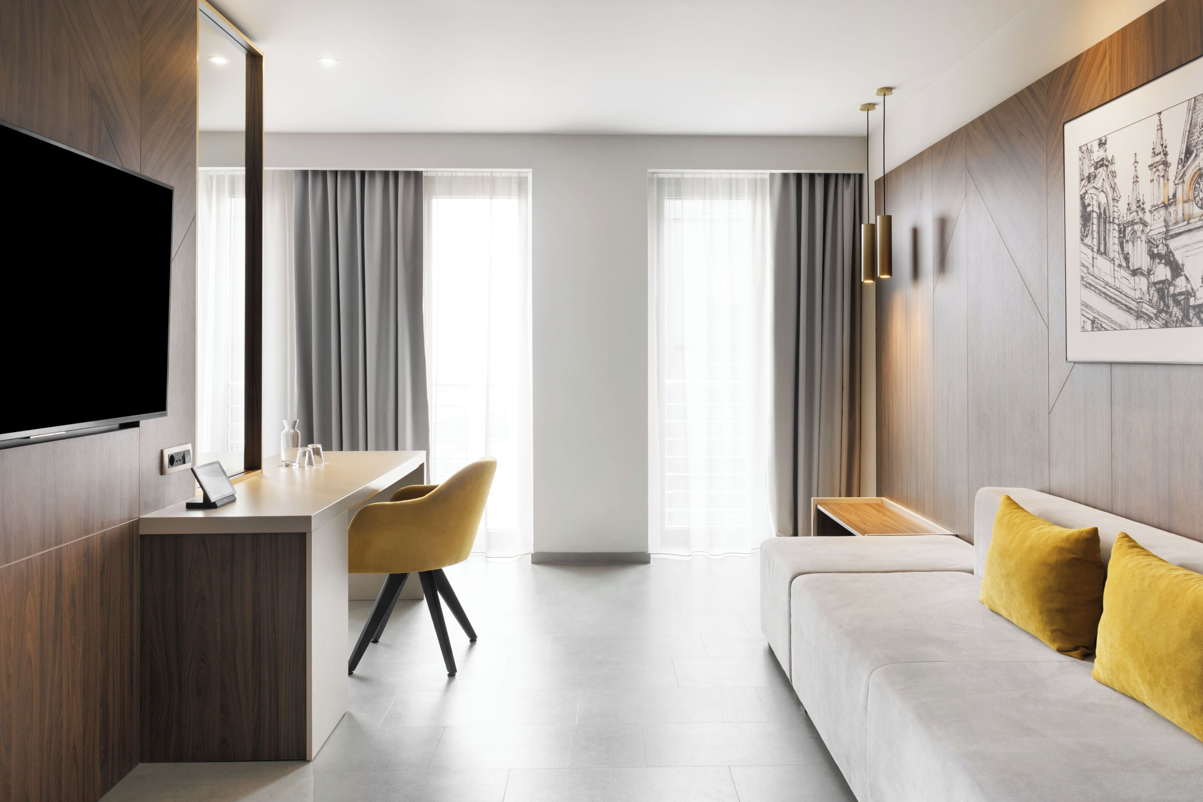 Quarto de hotel moderno com televisão de tela plana, escrivaninha com cadeira, sofá com almofadas e grandes janelas com cortinas.