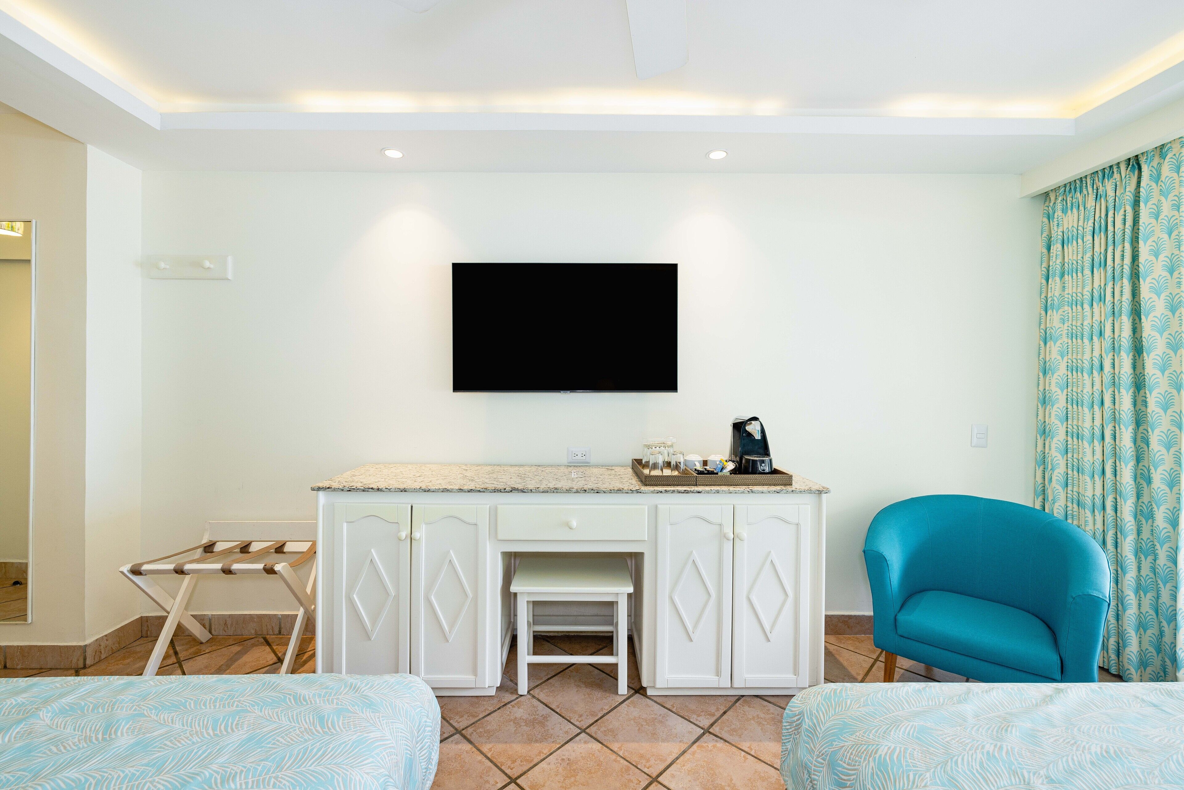 Quarto de hotel com cama, escrivaninha com cadeira, televisão e janela com cortinas.