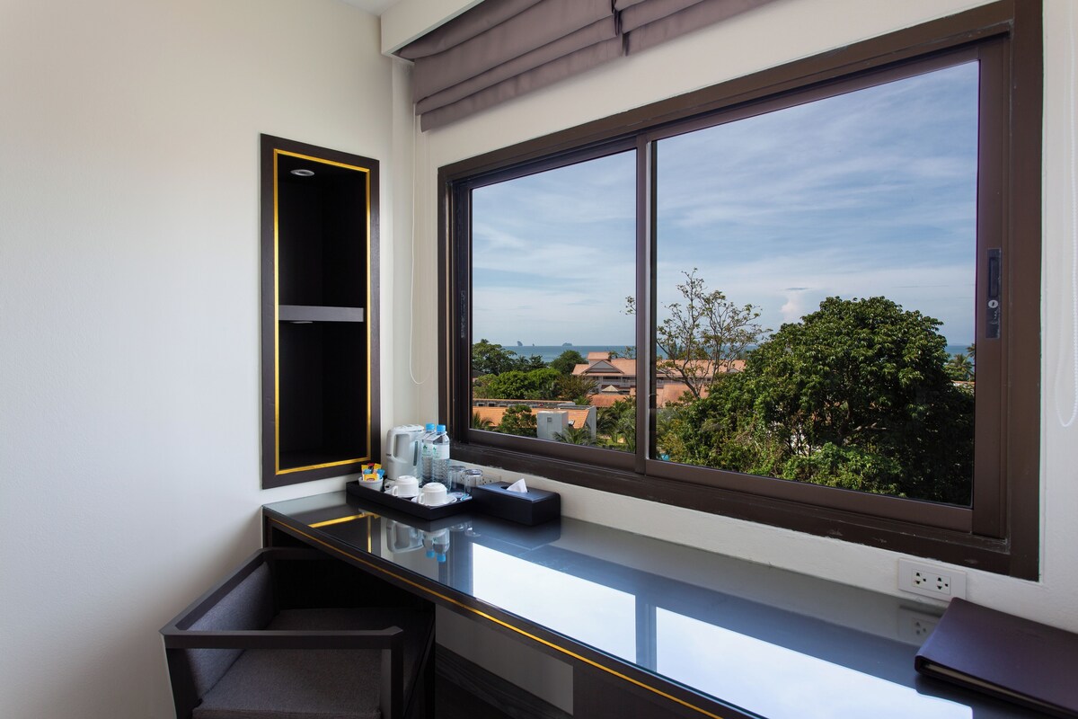 Quarto de hotel com uma grande janela, uma escrivaninha com um porta-cosméticos e um espelho na parede.