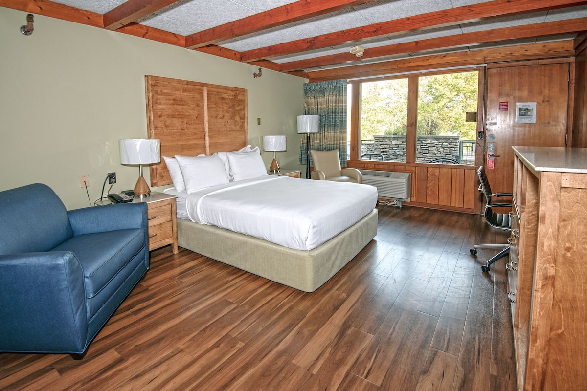 Quarto com cama, sofá azul, piso de madeira e janela com vista para área verde.
