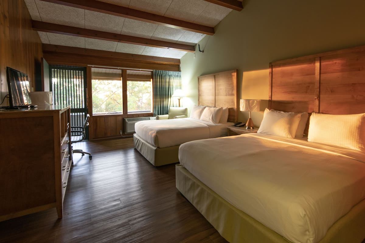 Quarto de hotel com duas camas, soalho em madeira, uma secretária com cadeira e janelas amplas com cortinas.