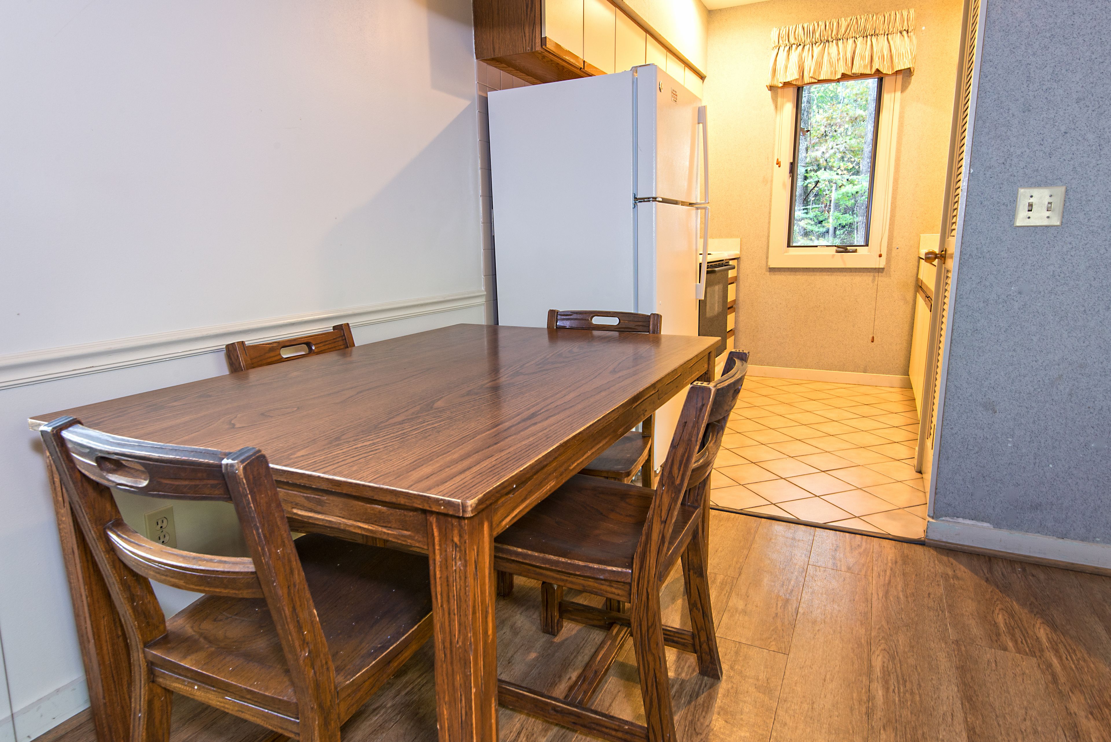 Uma área de refeições com mesa e cadeiras de madeira, geladeira branca e janela com cortinas.