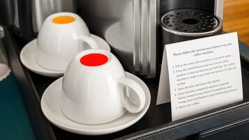 Máquina de café com instruções em um suporte, duas xícaras brancas com tampas vermelhas e uma chaleira prateada.