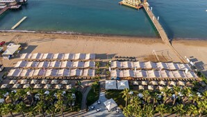 Vista aérea de um resort à beira-mar, com vários guarda-sóis, cadeiras de praia e um píer que se estende para a água.