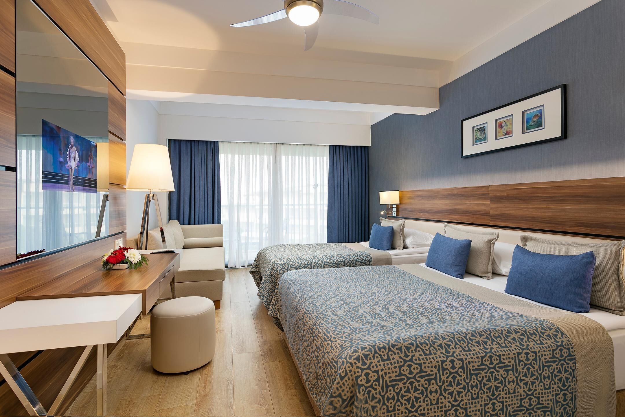 Quarto de hotel com cama, escrivaninha, cadeira, abajur, ventoinha no teto e janela com cortinas.