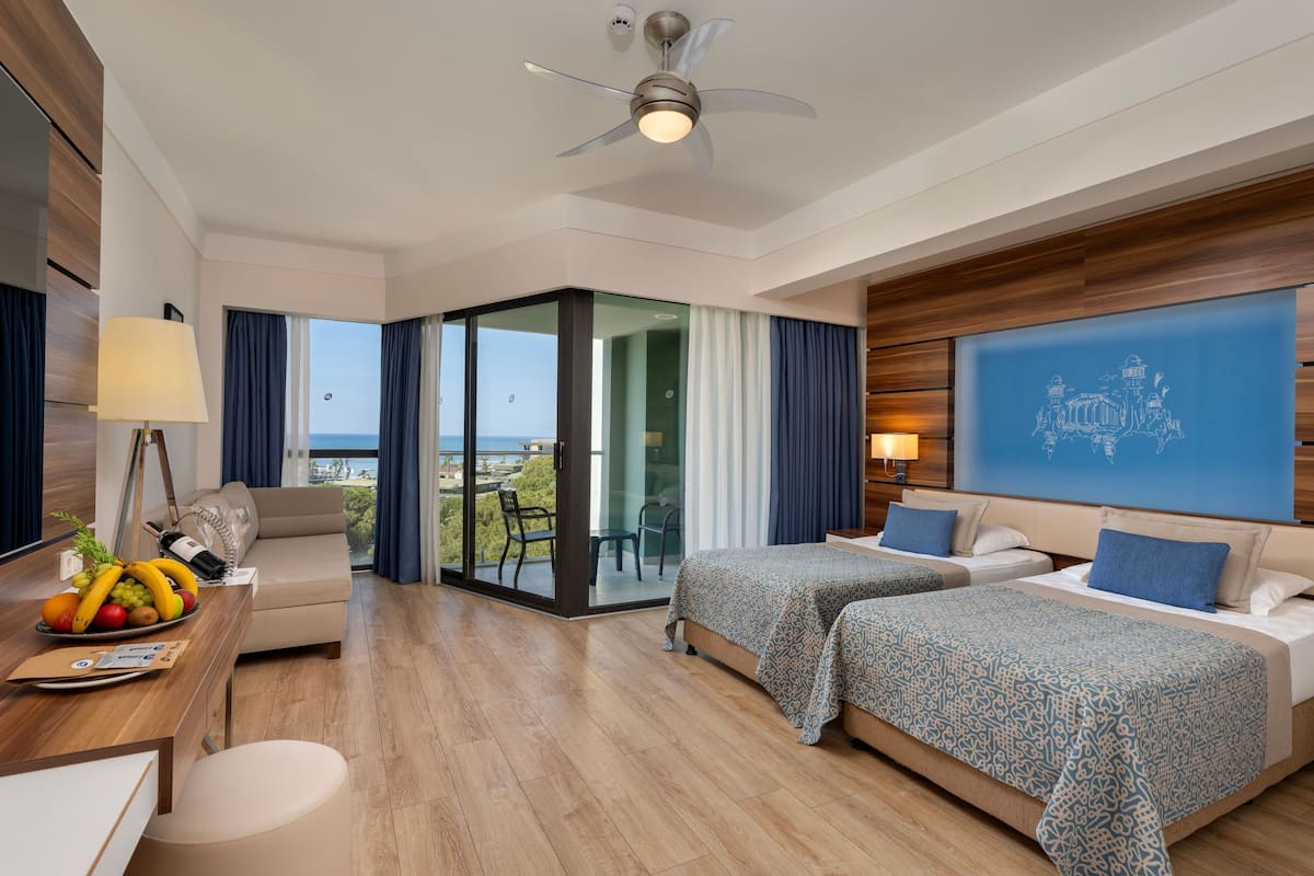 Quarto de hotel com varanda, duas camas, uma secretária com fruta e vista para o mar.