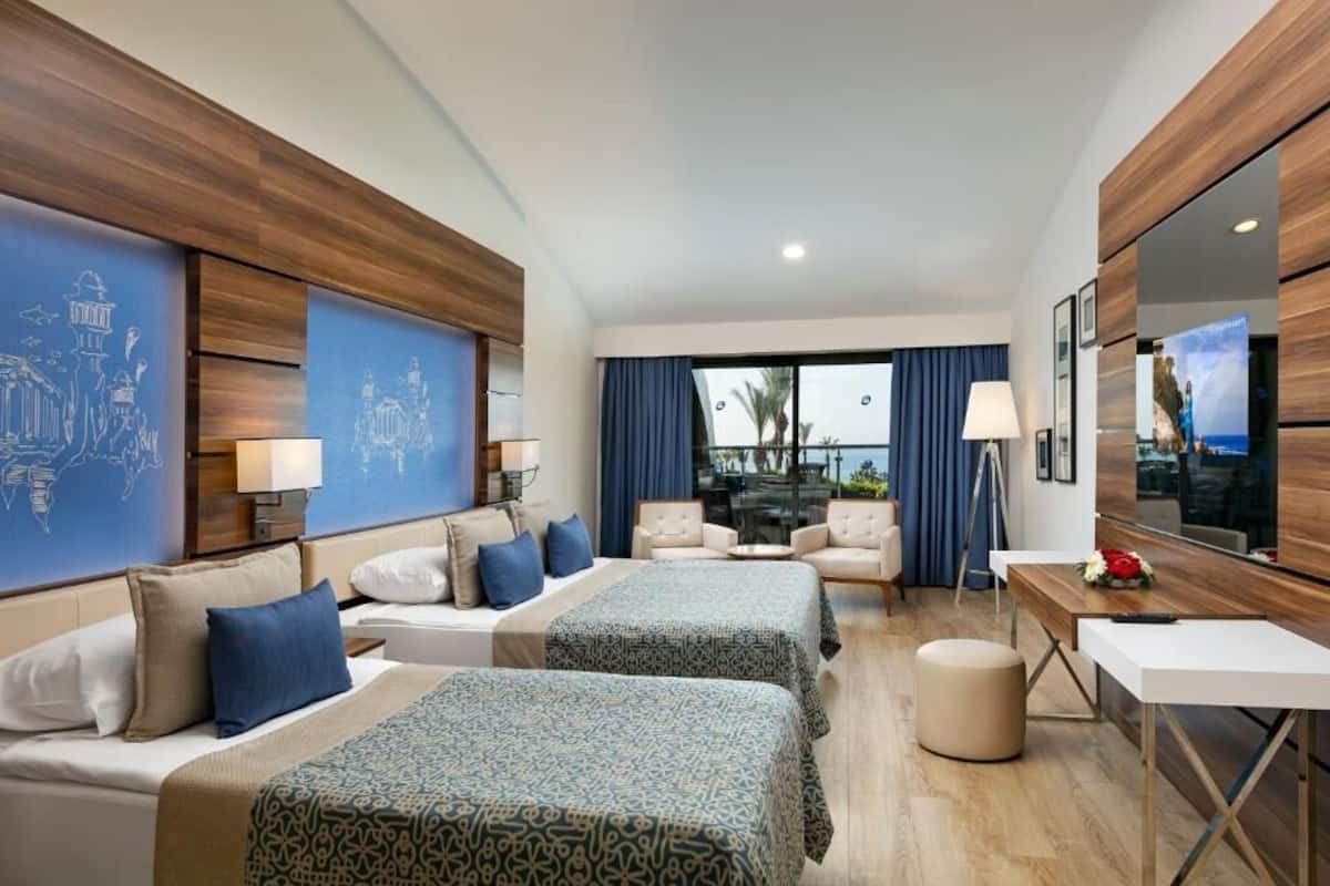 Quarto de hotel com duas camas, uma escrivaninha, uma cadeira, uma mesinha e uma janela grande com cortinas.